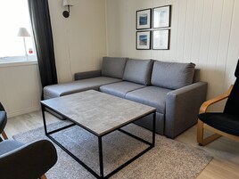 Uma sala de estar moderna com um sofá cinzento, uma mesinha e uma cadeira de madeira. Há quadros nas paredes e um abajur no parapeito da janela.