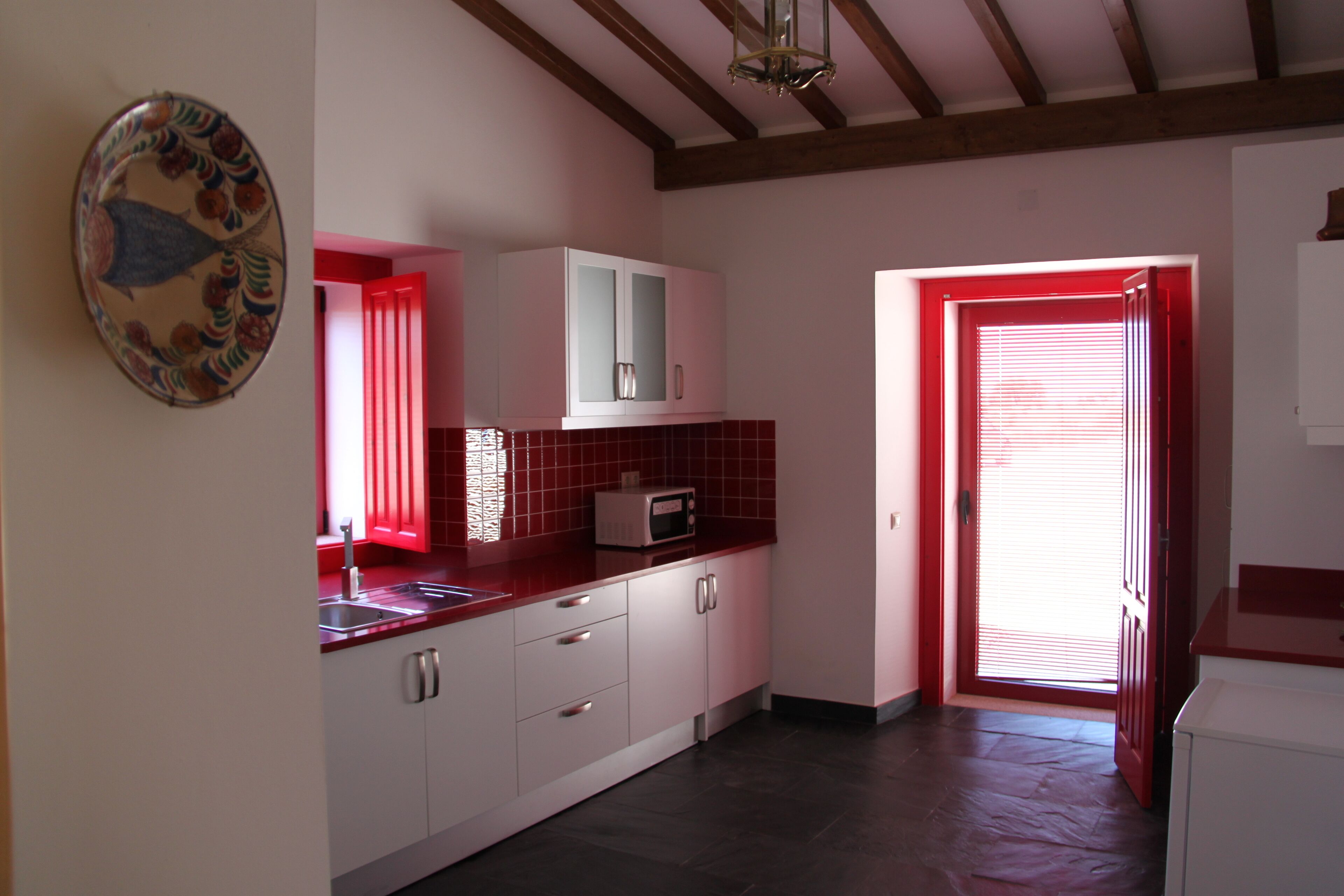 Cozinha com armários brancos, bancada vermelha, micro-ondas e porta com persianas vermelhas.