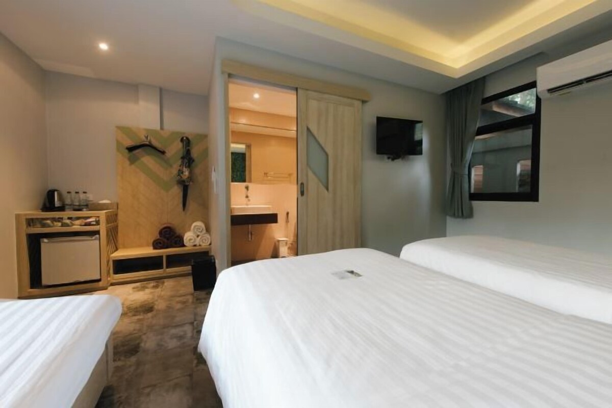 Quarto de hotel com duas camas, uma secretária pequena com cadeira, um espelho, televisão fixada na parede e área de chuveiro.