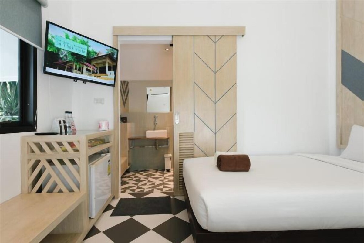 Quarto de hotel com cama, escrivaninha, televisão fixada na parede e banheiro visível por uma porta aberta.