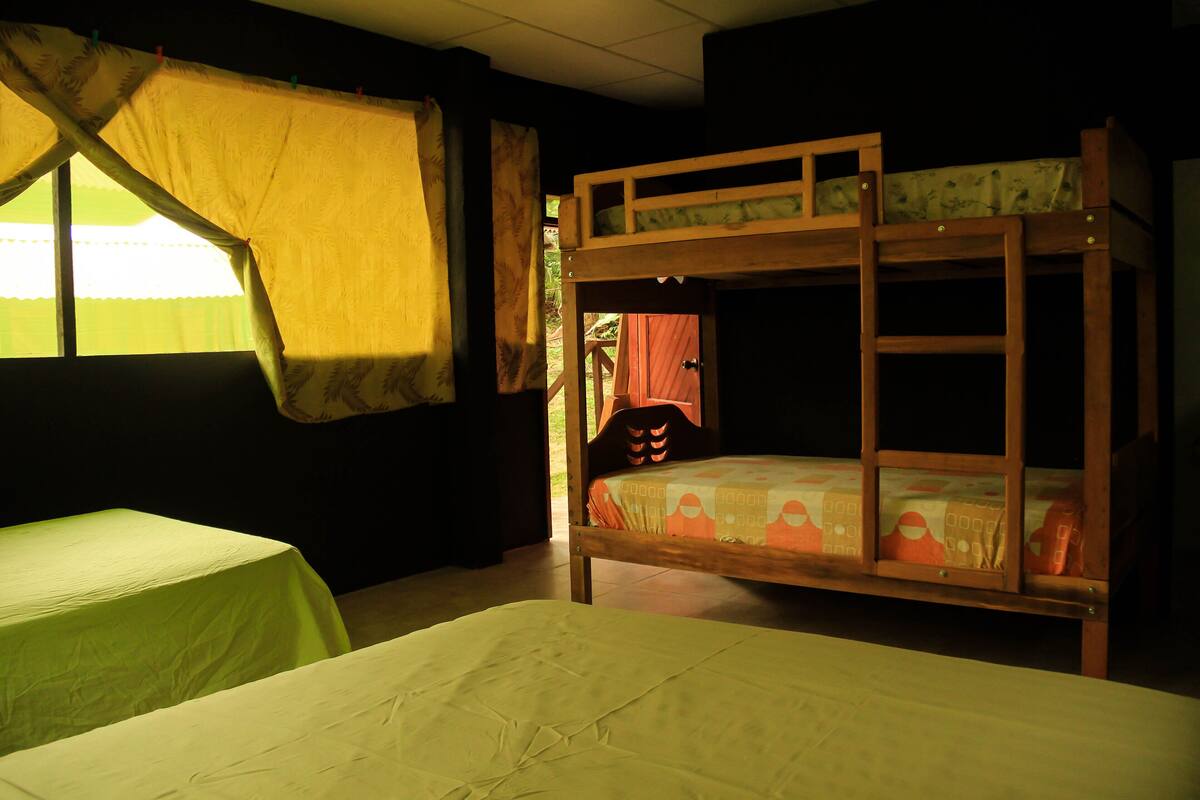 Quarto com beliche, janela e cortinas, uma mesa pequena e colcha verde.
