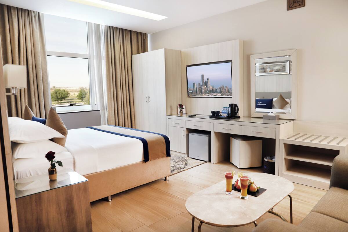Quarto de hotel com cama, escrivaninha, cadeira, mesa pequena com bebidas, televisão e vista para a cidade.