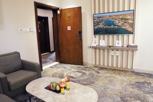 Um saguão de hotel com uma mesa de recepção, uma televisão exibindo uma vista urbana, uma área de estar com uma mesa e uma porta com número de quarto.