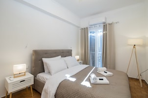 Quarto de hotel com uma cama, uma mesa de cabeceira com abajur, uma mesinha de cabeceira com gaveta e uma janela com cortinas.