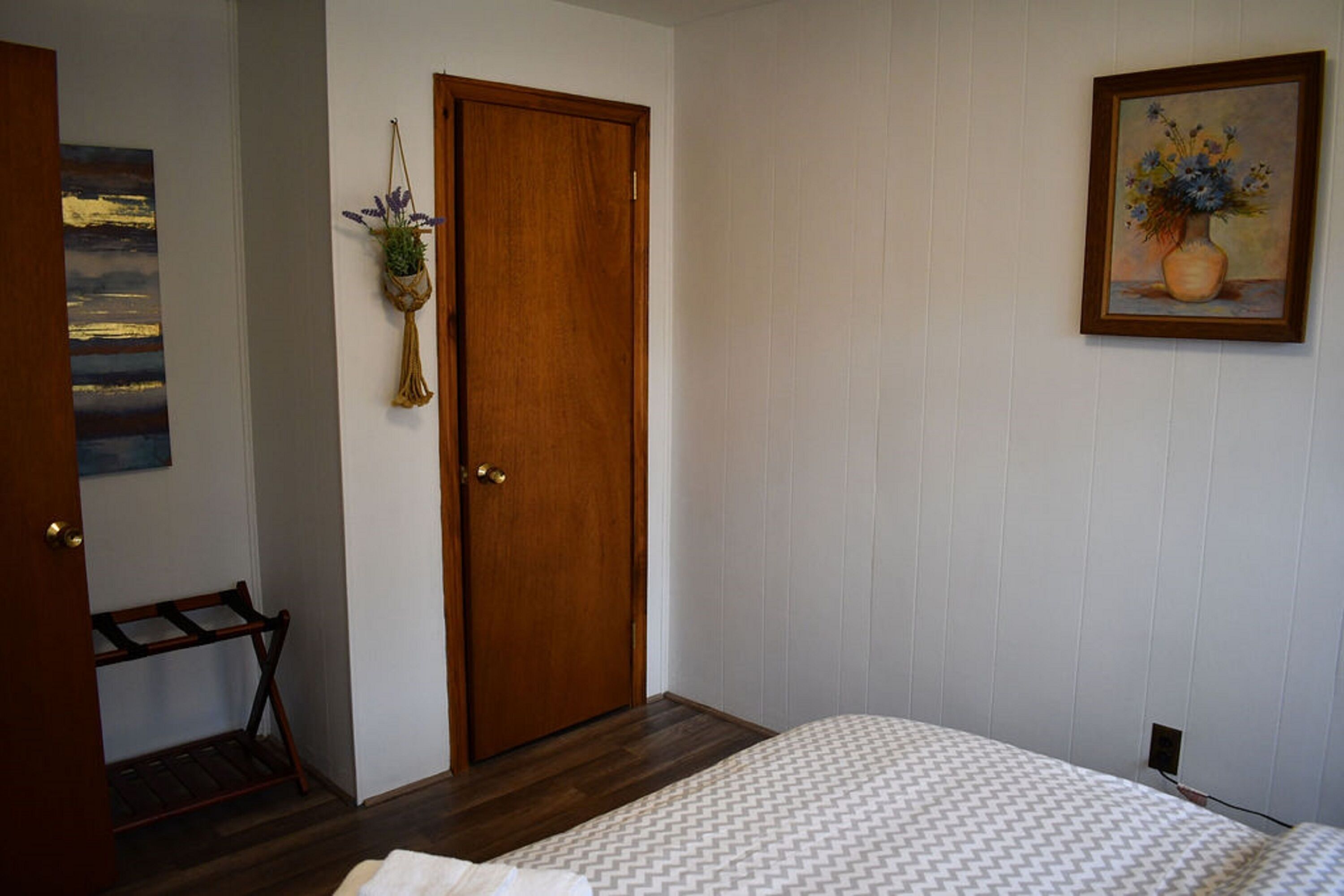 Quarto com porta de madeira, cama com colcha estampada, quadro na parede e planta pendurada.