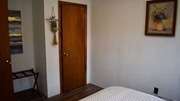 Quarto com porta de madeira, cama com colcha estampada, quadro na parede e planta pendurada.