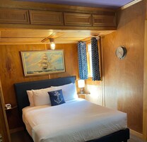 Cama bem arrumada com roupa branca, cabeceira escura e uma única almofada decorativa com desenho de ancora. Um quadro de um navio a vela está pendurado acima da cama, e um relógio de parede é visível à direita.