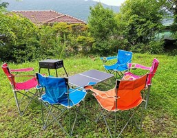 Uma mesa com um painel solar e um alto-falante portátil, cercada por cadeiras dobráveis coloridas em uma área externa gramada com árvores e uma casa ao fundo.