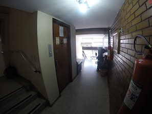 Um corredor estreito com um extintor do lado direito, uma porta com o aviso 'Não Fumar' e uma escadaria que leva a um andar superior.