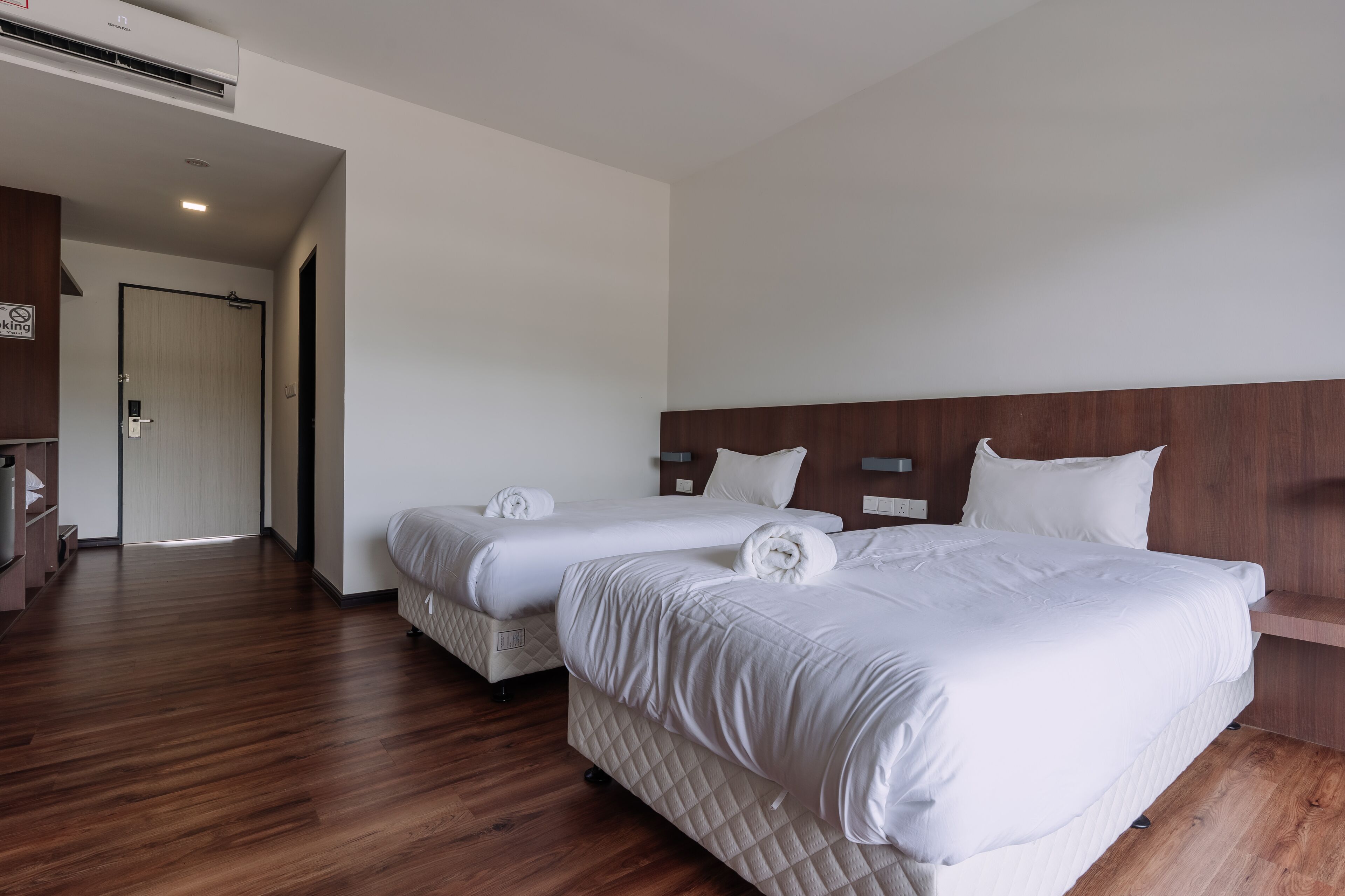 Quarto de hotel com duas camas, piso de madeira, cabeceira de madeira e parede branca.