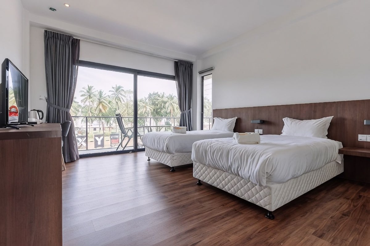 Quarto de hotel com duas camas, televisão, escrivaninha e varanda com vista para palmeiras.