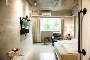 Quarto de hotel com cama, televisão na parede, mesa pequena, cadeira, luminária e janela com cortinas.