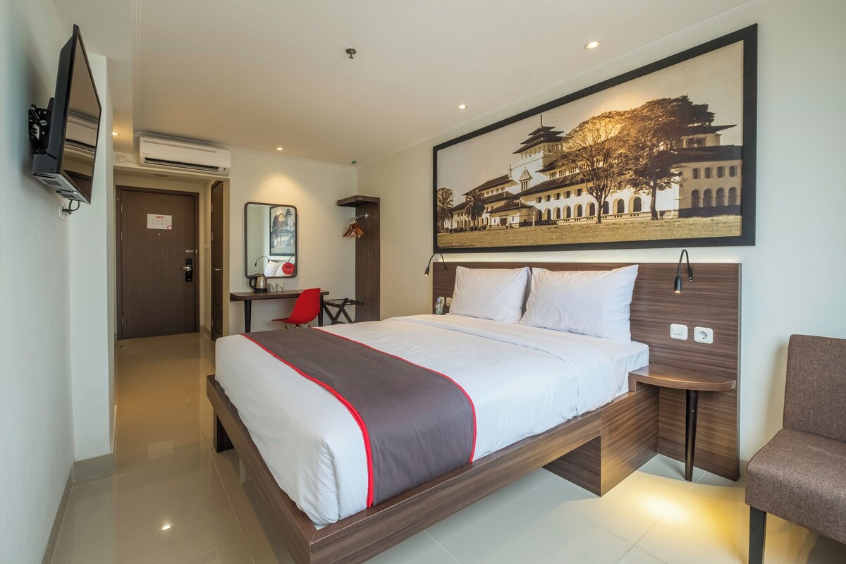 Quarto de hotel com cama, escrivaninha, cadeira e televisão fixada na parede.