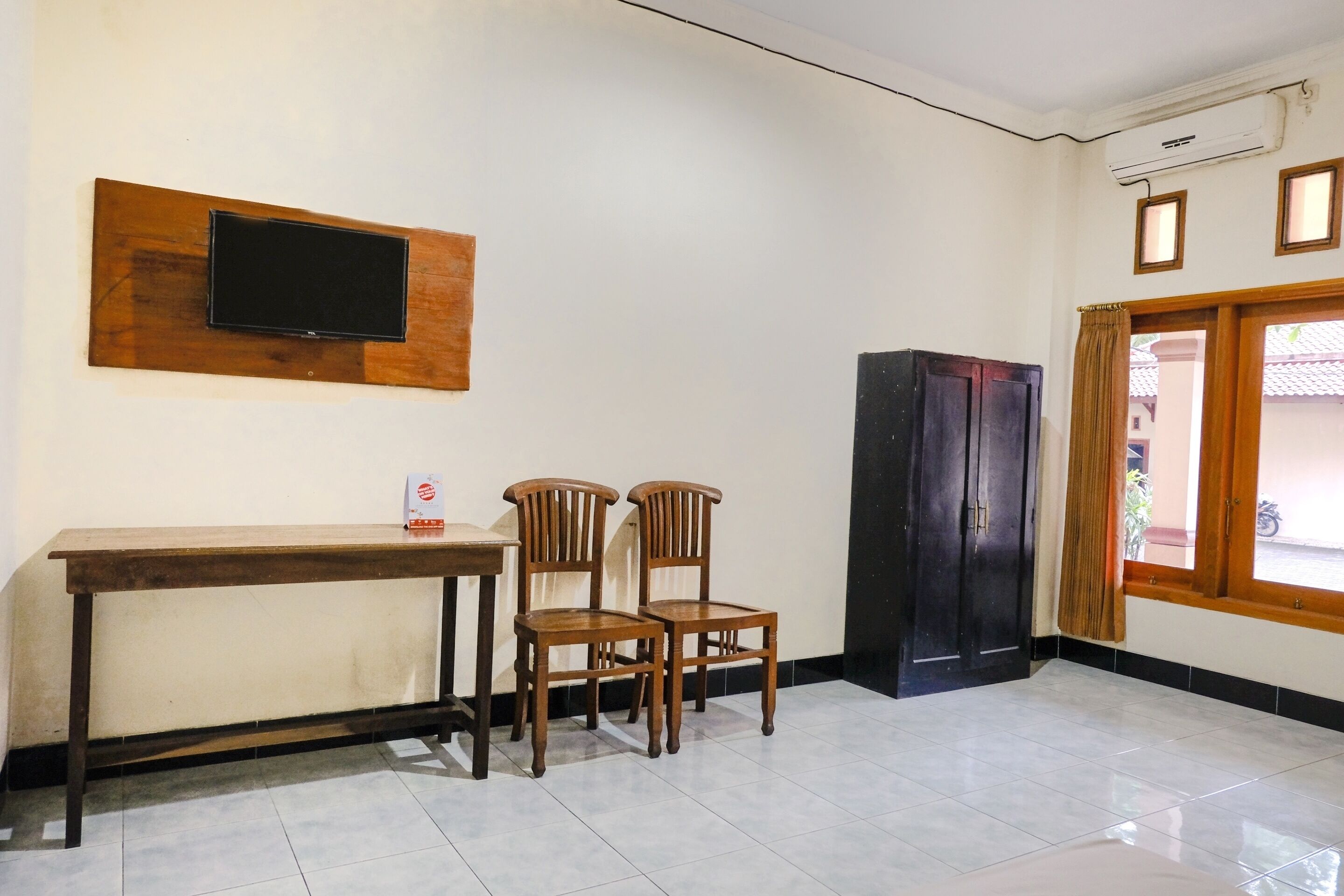 Quarto com mesa de madeira, duas cadeiras, televisão fixada na parede e um armário escuro.