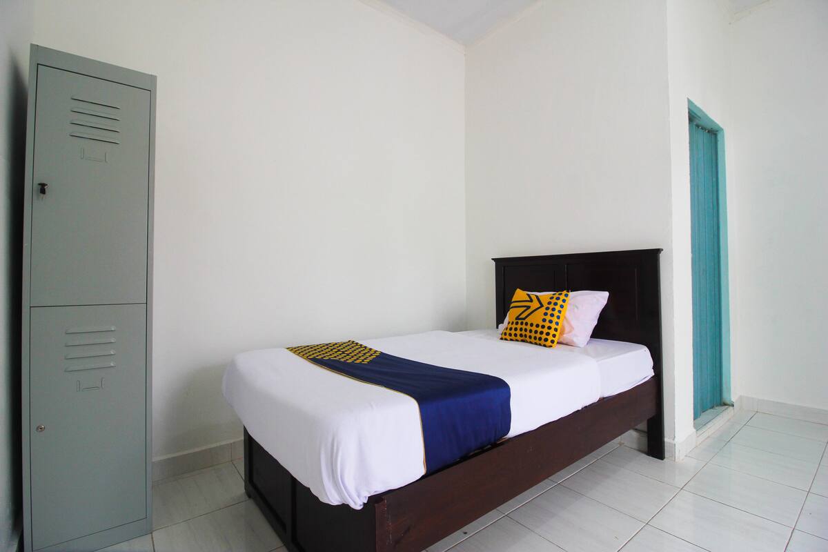Uma cama simples com colcha branca e azul, cabeceira de madeira e dois travesseiros com padrões geométricos. Um guarda-roupa de metal cinza está à esquerda da cama.