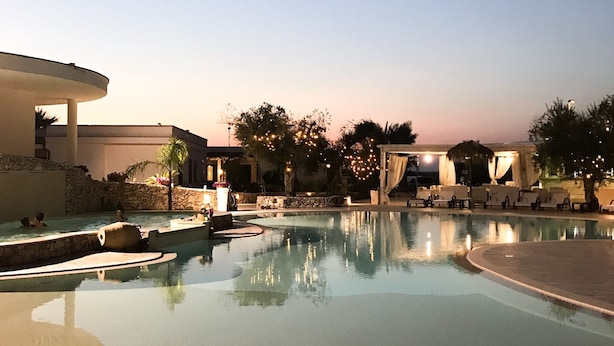 Uma área de piscina com céu claro, cadeiras de descanso e um edifício ao fundo.