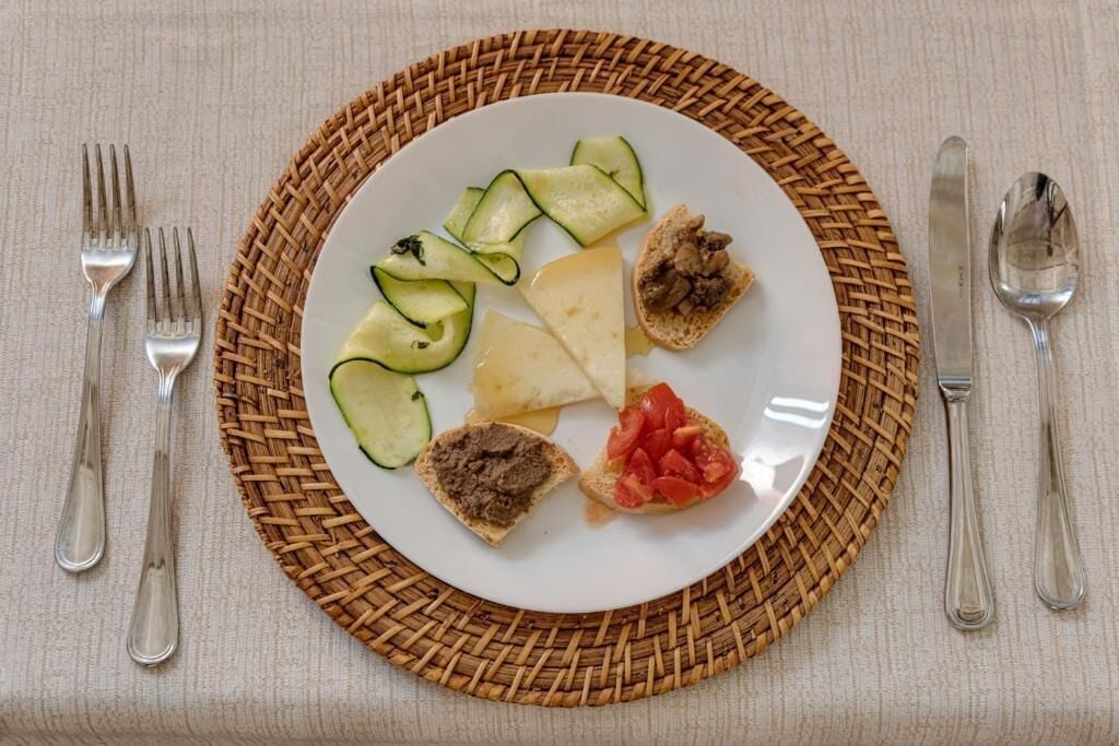 Prato com pepino fatiado, queijo e patê de carne no pão, acompanhado de garfo e colher sobre um tapete trançado.