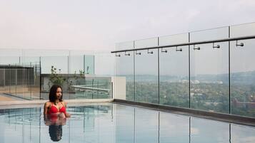 Mulher de fato de banho vermelho sentada em piscina com fundo de vidro transparente e vista para a cidade.