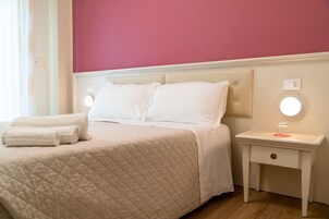 Cama bem arrumada com lençóis brancos e cobertura acolchoada, mesa de cabeceira com abajur e interruptor de luz na parede.