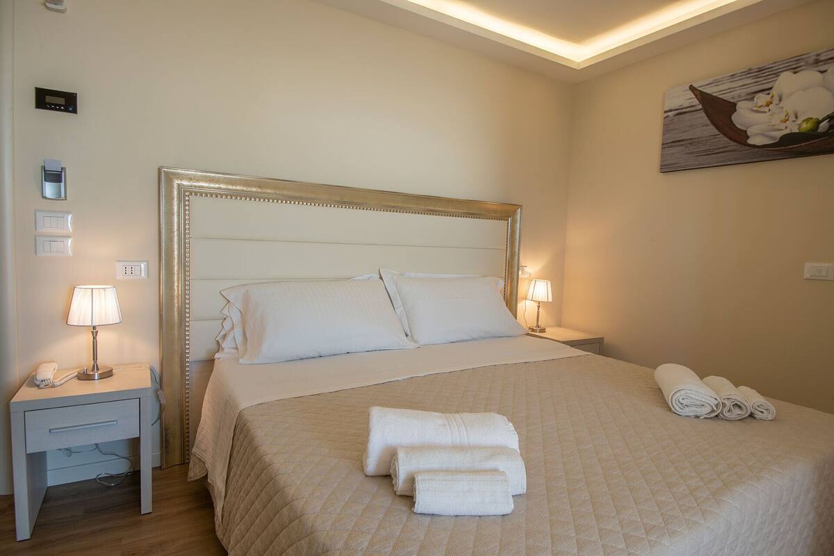 Uma cama bem arrumada com lençóis e toalhas brancas, uma mesa de cabeceira com abajur e um quadro na parede.
