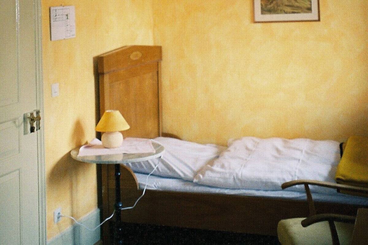 Quarto pequeno com uma cama de solteiro, cabeceira de madeira, mesa de cabeceira com abajur, um quadro na parede e uma porta com fechadura.