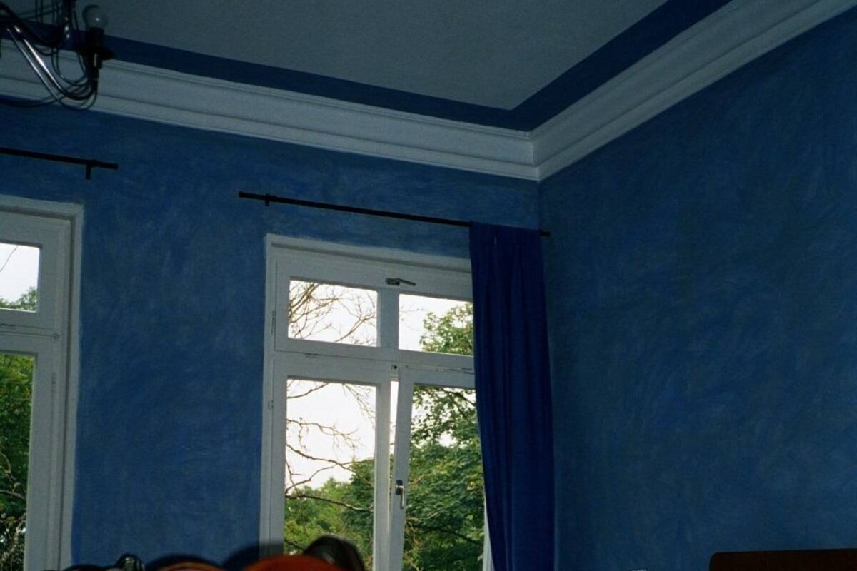 Quarto com cama, duas cadeiras, mesa de cabeceira, abajur e janela com cortinas.