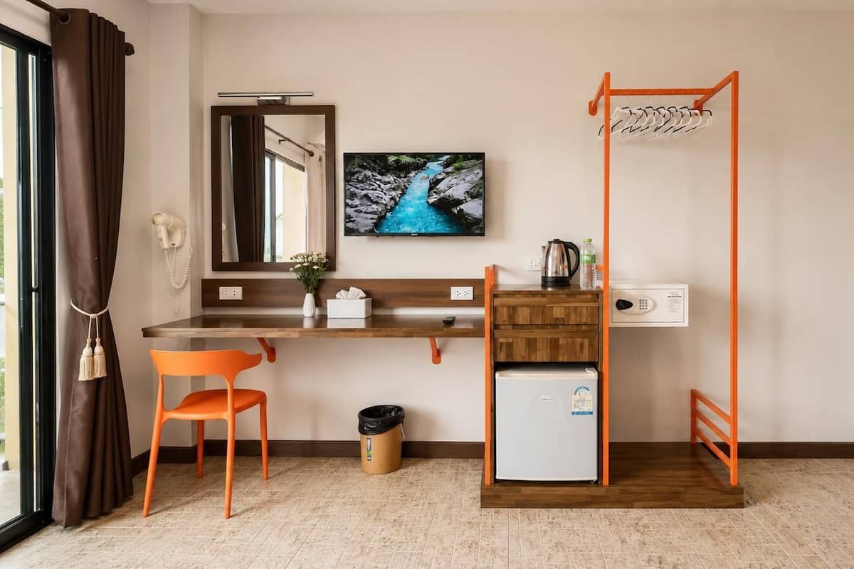 Quarto compacto com uma escrivaninha de madeira, uma cadeira, um refrigerador pequeno, uma TV e uma janela com cortinas.