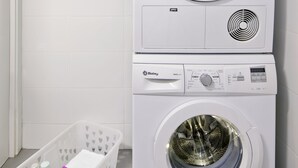 Máquina de lavar e secar sobrepostas numa lavanderia com parede de azulejos brancos e uma cesto de roupa com uma garrafa e uma caixa.