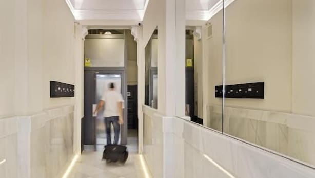 Corredor com uma pessoa caminhando em direção a um elevador, parede espelhada e chão de azulejos.