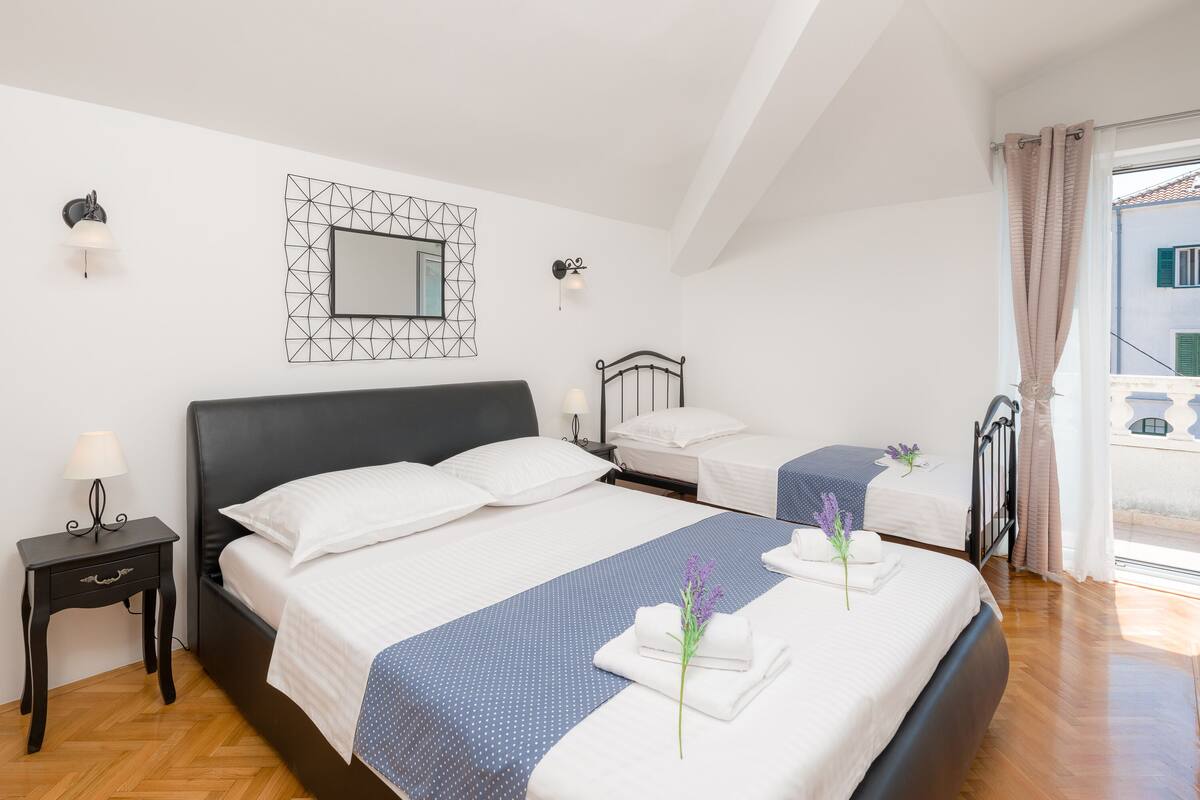 Quarto de hotel com uma cama grande, uma cama menor, mesa de cabeceira com abajur, espelho fixado na parede e janela com cortinas.