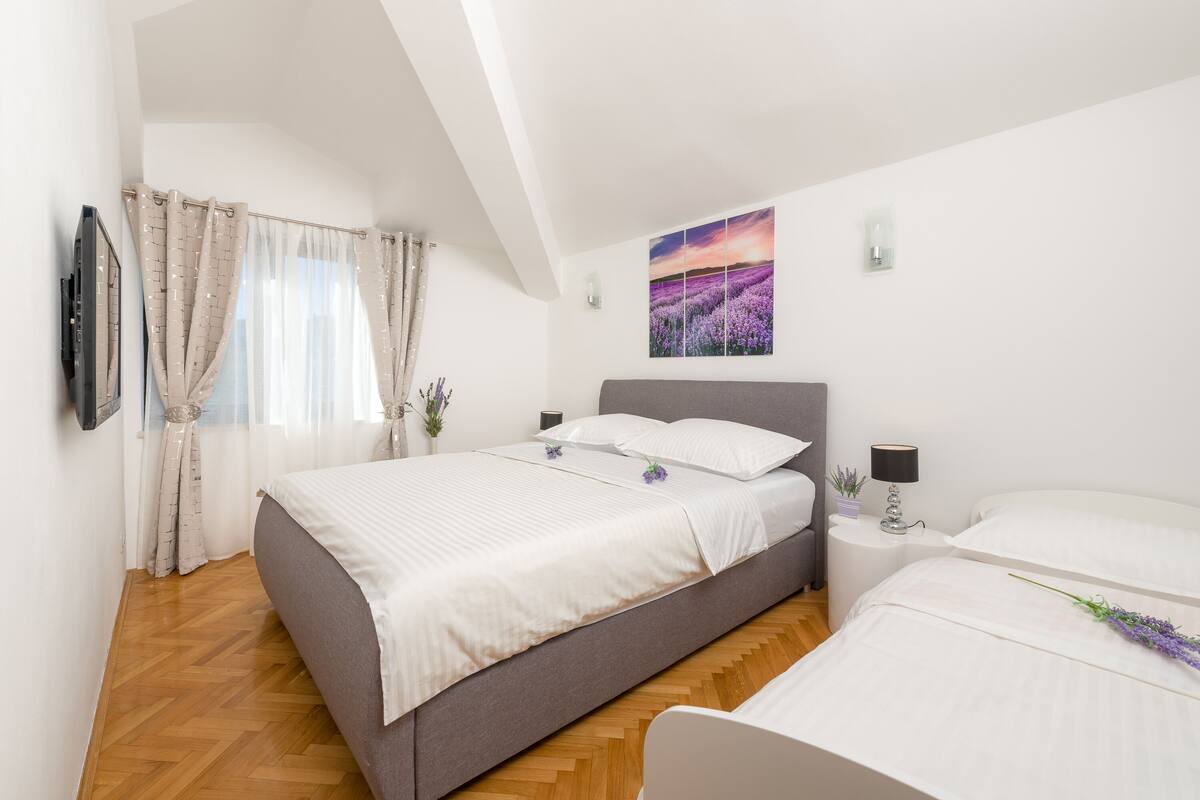 Quarto com cama, mesa de cabeceira, luminária, televisão na parede e janela com cortinas.