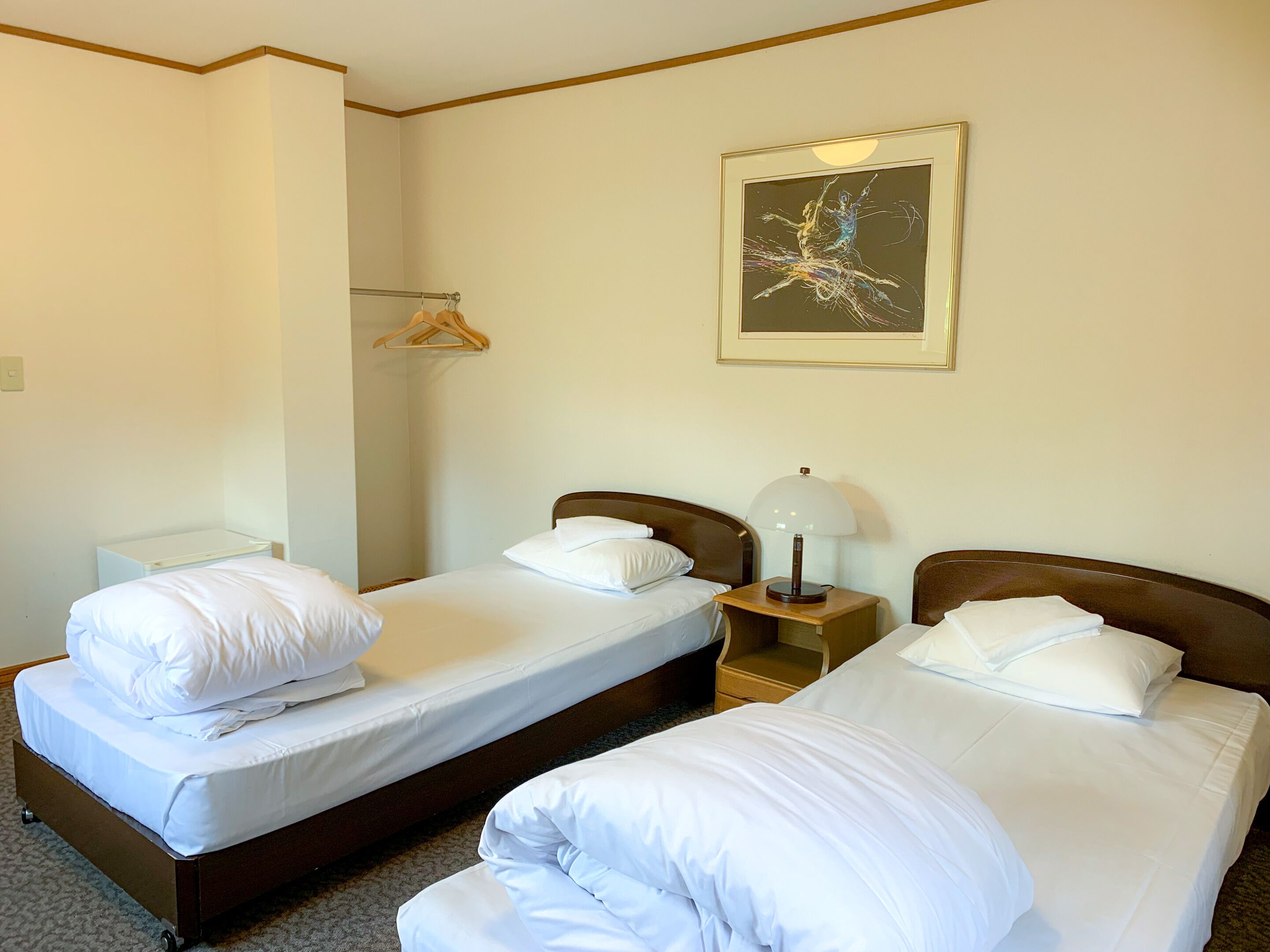 Quarto de hotel com duas camas de solteiro, uma mesa de cabeceira com luminária, um quadro abstrato emoldurado na parede e um cabideiro.