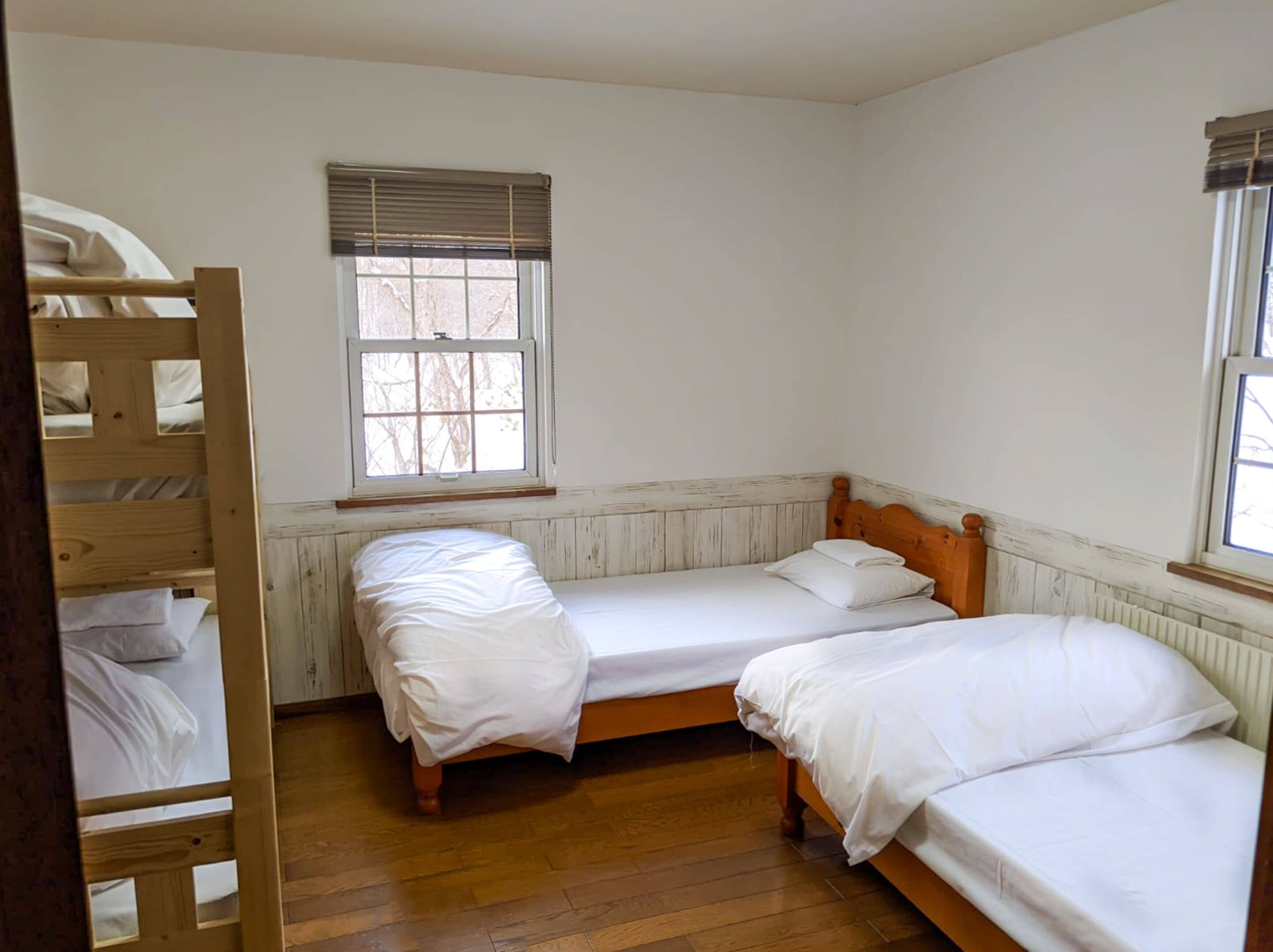 Quarto com duas camas de solteiro, uma beliche de madeira e uma janela com cortinas.