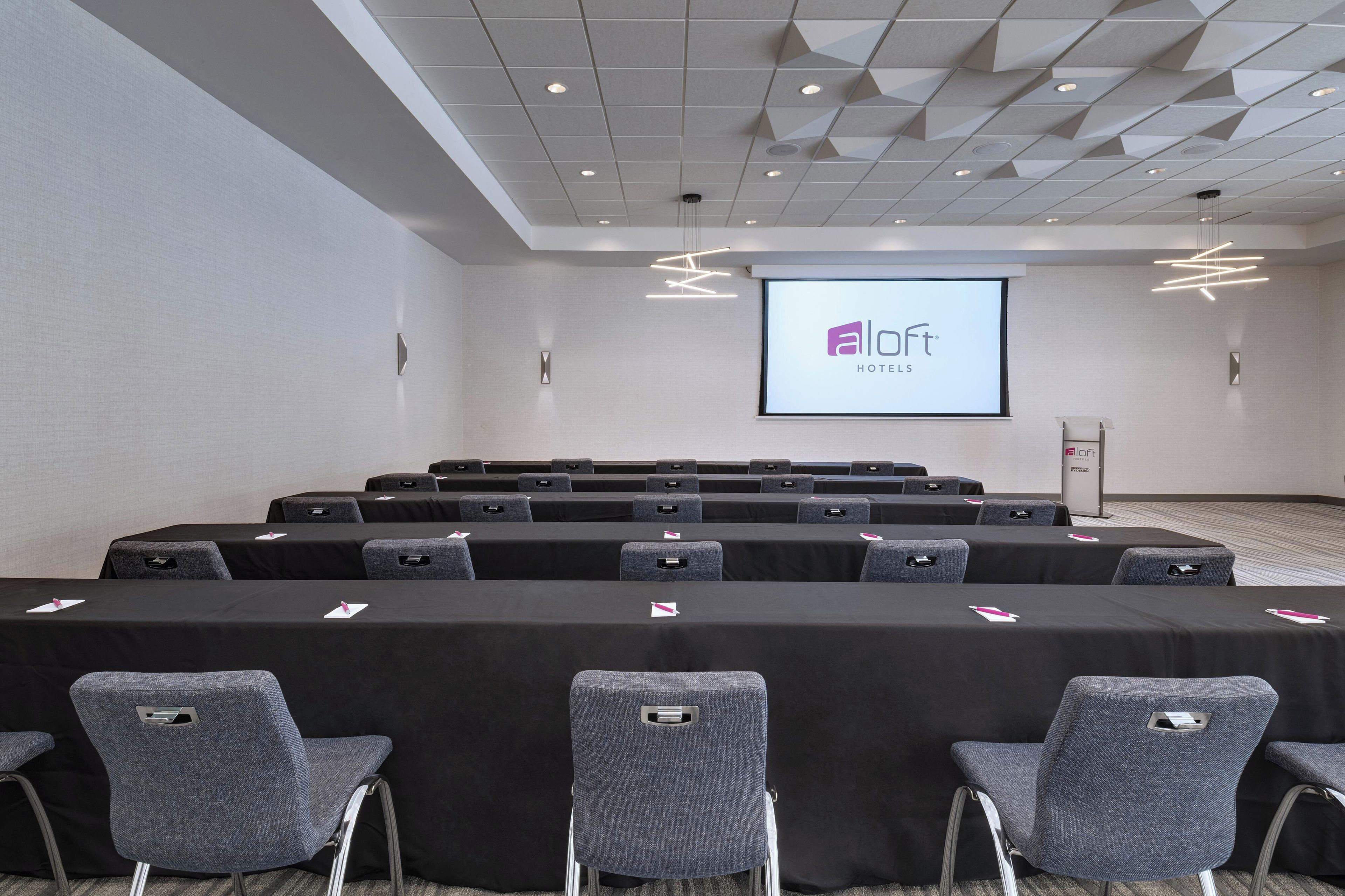 Sala de conferências com uma tela grande exibindo o logotipo de 'a loft hotels'.