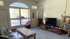 Uma sala de estar com sofá, televisão sobre um suporte de madeira, mesa com toalha estampada e janela com vista externa.
