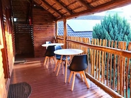 Um deck de madeira com mesas e cadeiras, um corrimão de madeira e vista para árvores e um edifício.