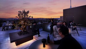 Um bar no terraço com mesas ao ar livre, uma grande árvore e vista para a cidade ao pôr do sol.