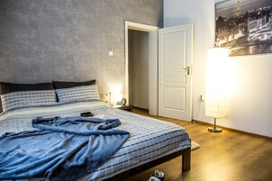 Quarto com cama, mesa de cabeceira, abajur e porta que dá para outro cômodo.