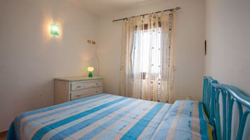 Quarto com cama, mesa de cabeceira, abajur e janela com cortinas.