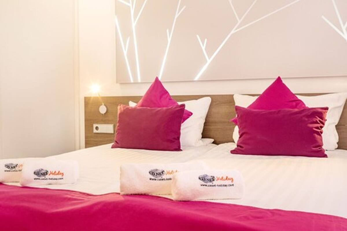 Quarto de hotel com cama, almofadas e luminária fixada na parede.