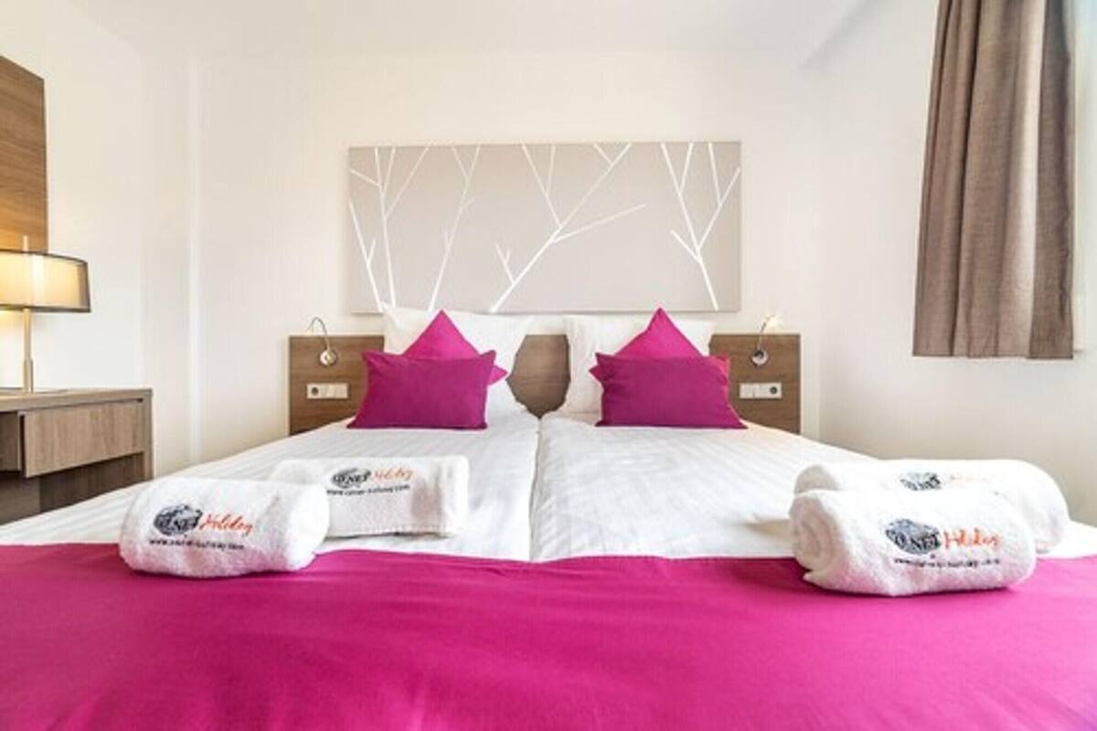 Quarto de hotel com cama, duas mesas de cabeceira, abajur, cabeceira com desenho de árvore e janela com cortinas.