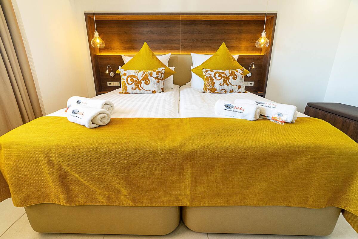 Uma cama de casal com colcha amarela e duas almofadas decorativas, cabeceira de madeira e duas luminárias pendentes.