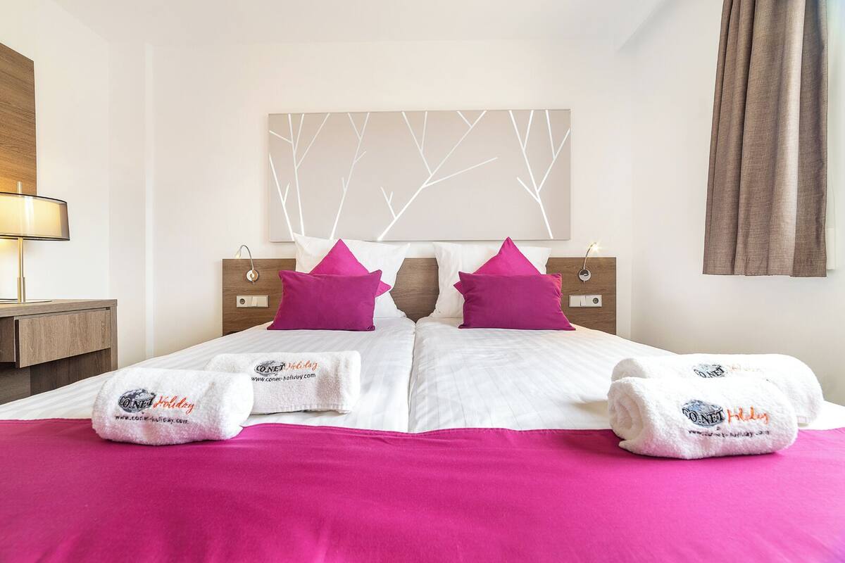 Quarto de hotel com uma cama grande, duas mesas de cabeceira, uma luminária, uma obra de arte na parede e uma janela com cortinas.