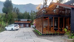 Um edifício moderno com área de estar coberta ao ar livre, um carro estacionado na frente e montanhas ao fundo.