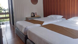 Quarto de hotel com duas camas, cabeceira de madeira, um ornamento circular na parede, uma pequena mesa com chaleira e garrafas, e vista para o verde através de uma porta deslizante.