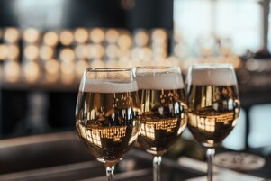 Três copos de cerveja numa bancada de bar, com luzes desfocadas ao fundo.