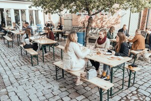 Um café ao ar livre com mesas e bancos de madeira, paredes de tijolo e uma árvore que oferece sombra.