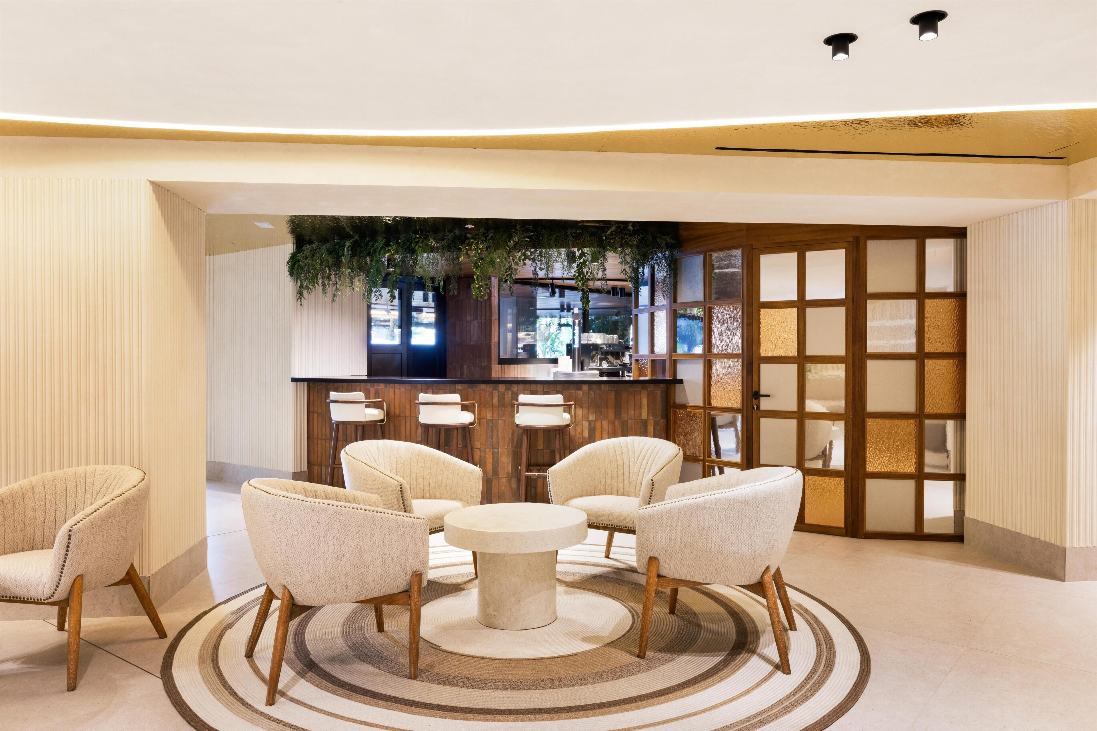 Um lobby moderno com uma mesa redonda e quatro cadeiras, uma área de bar com balcão de madeira e uma planta decorativa pendurada no teto.