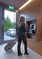 Mulher vestida para o trabalho utilizando um quiosque de autoatendimento no saguão de um hotel, com decoração em madeira e vista para um carro estacionado lá fora.