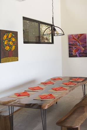Uma longa mesa de jantar com guardanapos e jogos americanos vermelhos, um lustre pendurado e duas pinturas nas paredes.